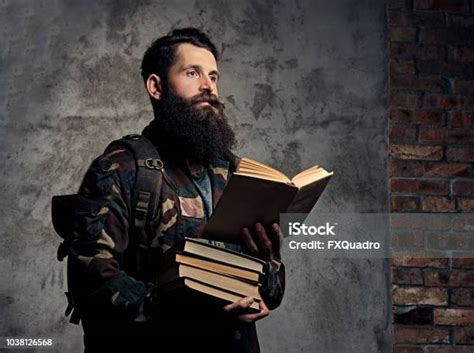 트렌디한 위장된 재킷을 입은 잘생긴 수염된 Hipster 어두운 배경에 고립 된 스튜디오에서 그의 손에 있는 책의 더미와 함께