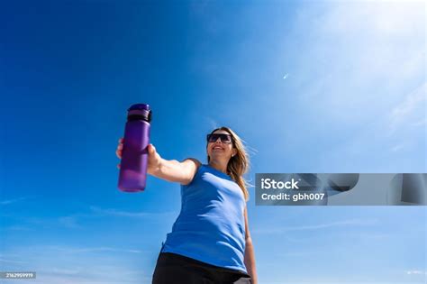 더운 여름에는 물을 마시는 데 주의합니다 푸른 하늘을 배경으로 물병을 주는 선글라스와 파란색 상의에 긴 금발 머리를 가진 젊은 현대 여성의 초상화 건강 에너지 생명의 기쁨