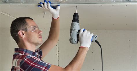 고글을 쓴 젊은 남자는 반짝이는 알루미늄 호일로 단열된 천장에 전기 드라이버를 사용하여 건식 벽체 매달린 천장을 금속 프레임에 고정합니다 리노베이션 건설 직접 개념을