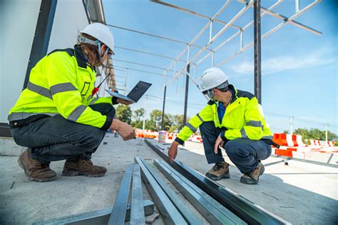 눈에 잘 띄는 재킷과 헬멧을 쓴 두 명의 엔지니어가 모듈식 건축 현장에서 노트북을 사용하여 건설 세부 사항에 대해 논의하며 강철 빔에 초점을 맞추고 정확성 팀워크 및 현장