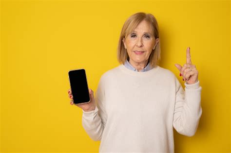 노란 배경 위에 고립된 서 손가락을 가리키는 휴대 전화 화면을 보여주는 흰색 스웨터를 입은 노인 미소 가리키기에 대한 스톡 사진