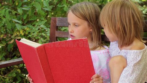 Older Sister Reading A Book Aloud To Her Younger Sibling On A Wooden