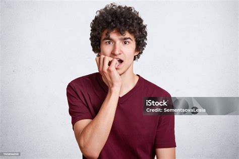 어두운 곱슬 머리를 가진 젊은 남자의 스튜디오 샷 자연스럽 게 물고 손톱과 흰색 배경에 서 있는 동안 카메라를 직접 보고 보고
