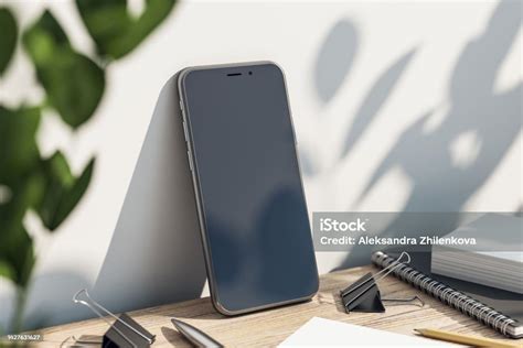 현대 스마트 폰의 빈 검은 색 화면에 대한 원근감보기는 사무용 도구가있는 나무 테이블에 텍스트 또는 로고를위한 카피 스페이스와 햇볕이 잘 드는 흰색 벽 배경에 녹색 식물이