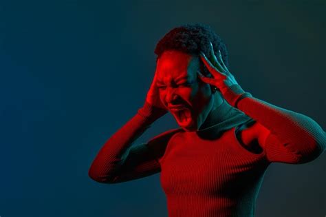어두운 스튜디오 배경 위에 네온 불빛에 손을 잡고 머리를 잡고 미친 듯이 소리치는 감정적인 젊은 여성의 헤드샷 초상화 프리미엄 사진