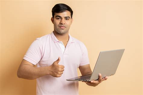 캐주얼한 옷을 입은 젊은 인도 남성이 노트북을 손에 들고 베이지색 스튜디오 배경에 고립되어 엄지손가락을 치켜들고 있다 사람들의
