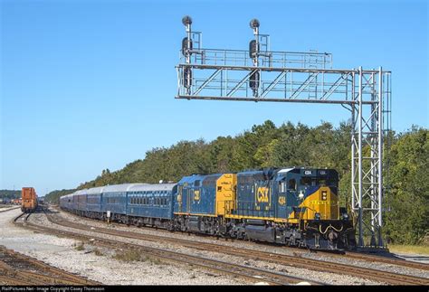 A Blue And Yellow Train Traveling Down Tracks Next To Some Trees On A Sunny Day