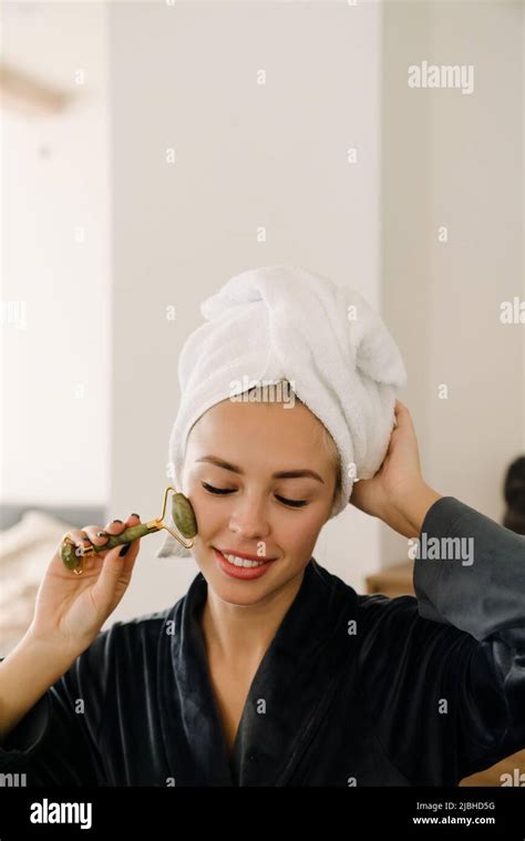 White Woman Wrapped Towel Massaging Face With Jade Roller At Home Stock
