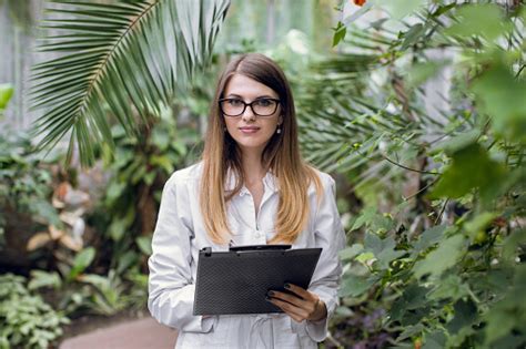 온실 과 열대 식물 온실에서 자라는 식물을 검사하고 클립 보드를 들고 메모를 만드는 동안 젊은 매력적인 백인 여성 과학자가 카메라에 포즈를 취했습니다 Stem 주제에 대한