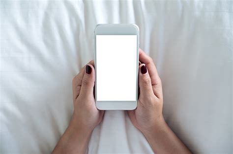 하얀 침대에 앉아 텅 빈 데스크탑 스크린으로 흰색 휴대 전화를 들고 여자의 손의 상단 보기 이랑 이미지 형판에 대한 스톡 사진