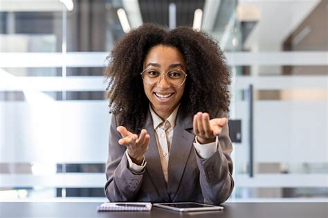 젊은 아프리카계 미국인 여성이 사무실의 책상에 앉아 원격으로 카메라와 대화하고 미소를 지으며 손으로 몸짓하는 클로즈업 초상화 가상현실에 대한 스톡 사진 및 기타 이미지