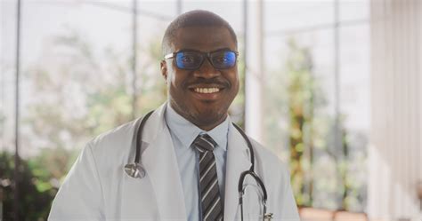 카메라를 위해 포즈를 취하고 미소를 짓고 카메라를 바라보는 흑인 의료 전문가의 초상화 안경을 쓰고 흰 코트를 입은 아프리카
