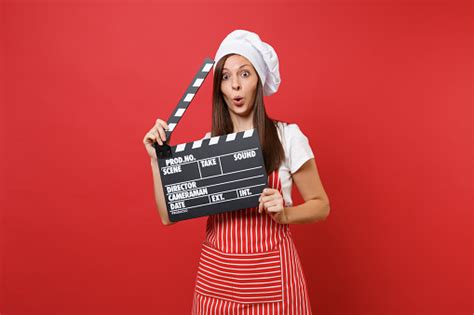 주부 여성 요리사 요리사 또는 줄무늬 앞치마 흰색 티셔츠 모자 요리사 모자 빨간 벽 배경에 고립 된 베이커 여자 만들기 Clapperboard 고전적인 흑인 영화를 들고