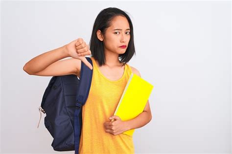 화가 얼굴 엄지 손가락으로 싫어하는 것을 보여주는 부정적인 기호 거부 개념으로 고립 된 흰색 배경 위에 책을 들고 배낭을 입고 젊은 중국 학생 여성 가방에 대한 스톡 사진