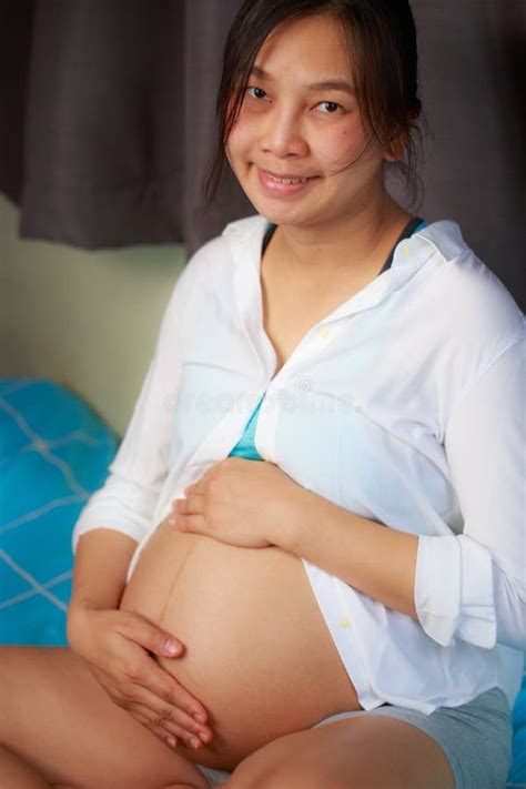 Pregnant Woman Smiling While Sitting On Bed Showcasing Her Belly Stock
