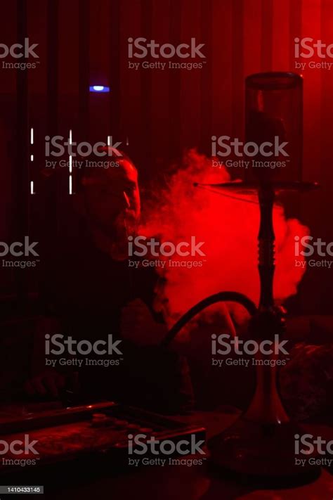 한 젊은이가 어두운 물 담뱃대 방에서 물 담뱃대를 피우고 붉은 빛이 그들에게 비춥니다 25 29세에 대한 스톡 사진 및 기타