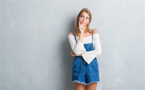 손으로 턱 질문 잠겨있는 표현에 대 한 생각에 그런 지 회색 벽에 서 있는 아름 다운 젊은 여자 사려깊은 얼굴 미소 의심