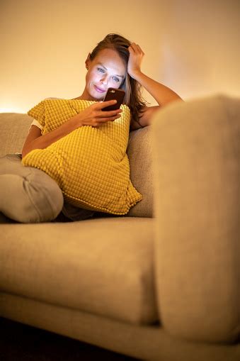 그녀의 휴대 전화를 사용 하 여 그녀의 현대적인 아파트에는 편안한 소파에 귀여운 젊은 여자 거실에 대한 스톡 사진 및 기타 이미지 거실 건강하지 못한 생활방식 말하기