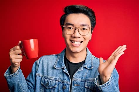 고립된 빨간 배경 위에 커피 한 잔을 마시는 젊은 잘생긴 중국 남자는 매우 행복하고 흥분 큰 미소와 함께 비명을 지르는 승리를