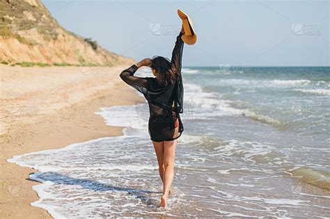 暑假。美丽时尚的女人戴着帽子走在沙滩上的海浪和放松。适合年轻女子，浅黑色衬衫和草帽享受热带岛屿度假。