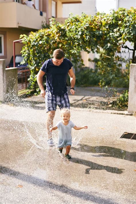 Dad And Daughter Run Through The Puddles Raising A Fan Of Spray Stock