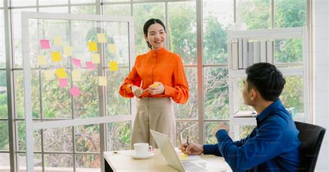 비즈니스 사람들은 사무실의 프로젝트 계획 보드에서 일하고 동료 친구와 대화하여 프로젝트 개발을 분석합니다 그들은 유리 벽에 게시 된 스티커 메모를 사용하여 정리합니다