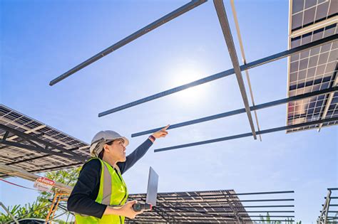 안전 헬멧과 반사 조끼를 입은 기술자가 노트북을 사용하여 태양 전지판 구조를 검사하고 있습니다 태양 전지판은 맑고 푸른 하늘 아래 강철 간이 차고 프레임워크에 장착됩니다