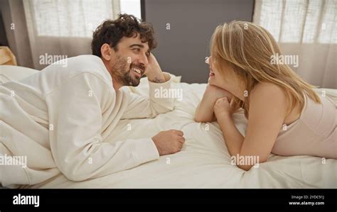 A Smiling Man And Woman Lying On A Bed Facing Each Other In A Cozy