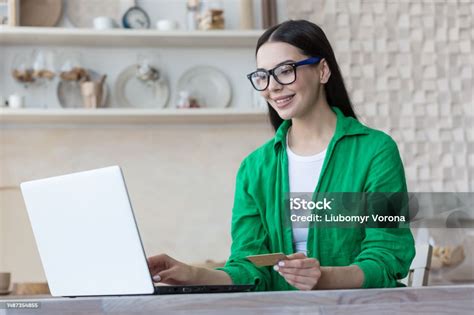 Молодая Женщина Сидит На Кухне Дома За Столом С Ноутбуком Держит Кредитную Карту И Занимается