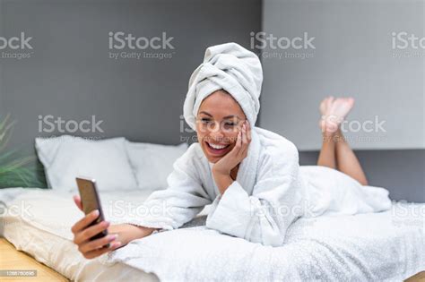 호텔 방에 침대에 수건을 들고 있는 젊은 아름다운 여성 흰색 테리 목욕 가운을 입고 휴대 전화로 셀카를 찍고 그녀의 얼굴에