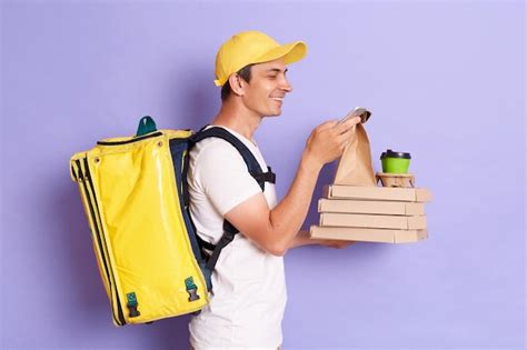 黄色い帽子をかぶった配達員と、携帯電話を使って紫色の背景に隔離された段ボールのフラットボックスにピザを入れたサーモバックパックを持ったtシャツ