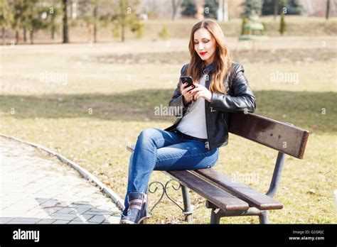 Young Smiling Woman Using Smartphone Sitting On Bench In Park
