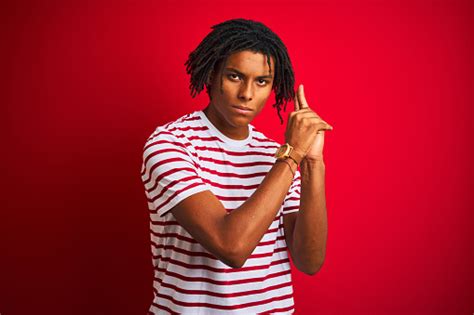 고립 된 빨간 배경 위에 서있는 줄무늬 티셔츠를 입은 공포를 가진 젊은 아프로 남자손 제스처로 상징적 인 총을 들고 살인 사격 무기 화가 난 얼굴 T 셔츠에 대한 스톡 사진