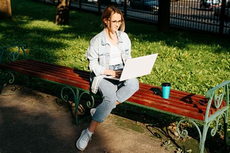 평범한 옷과 노트북에서 일하는 안경을 쓰고 있는 젊은 미소 짓는 여성이 일회용 커피 한 잔을 들고 공원 벤치에 앉아 인터넷을 검색합니다 프리랜서 원격 작업 사무실 아웃도어