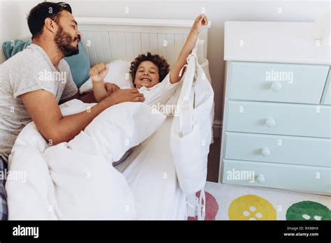Man Waking Up His Son Lying On Bed Father Lying Beside His Son On Bed And Talking To Him Stock