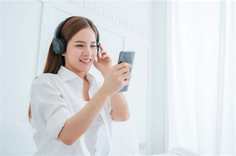 아름 다운 매력적인 아시아 여자 헤드폰 손 홀드 스마트 폰 흰색 침실 배경 초상화 아시아 긴 머리 흰색 셔츠 여자에서 노래 듣기