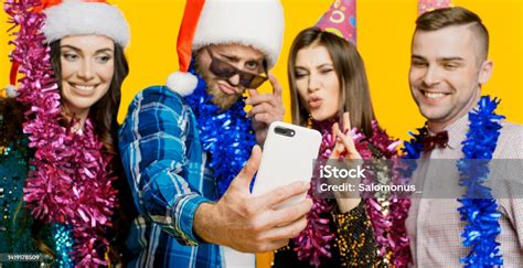 파티 모자와 산타 클로스 모자를 쓰고 틴젤을 착용 한 20 대 청년들은 비디오를 촬영하거나 소셜 미디어 용 스마트 폰에서 셀카를 찍습니다 손에 스마트폰에 선택적으로 초점을