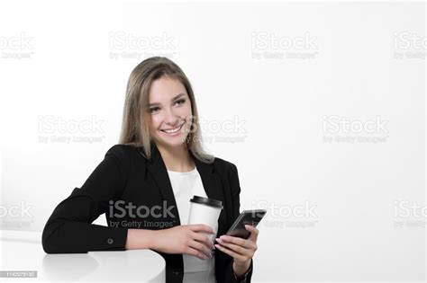 검은 양복과 흰색 블라우스에 젊은 아름 다운 여자는 사무실 테이블에 앉아 있다 흰 종이 컵에서 커피의 뜨거운 음료를 마신다 스마트 폰에 메시지를 쓰고 통신 하 고 미소를