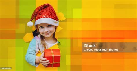 빨간 산타 모자를 쓴 패니 리틀 미소 소녀는 여러 가지 색의 종이에 구멍을 내다보고 선물이 있는 빨간 상자를 손에 들고 있다 크리스마스와 새해 개념입니다 복사 공간 팝 아트