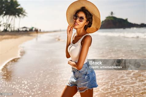 밀짚모자를 쓰고 선글라스를 낀 젊은 아프리카 여성 모델이 일출에 바닷가에서 포즈를 취하고 있다 새벽에 경치 좋은 바위 같은 녹색 섬과 바다 조수에 반대하는 흑인 여성 다인종