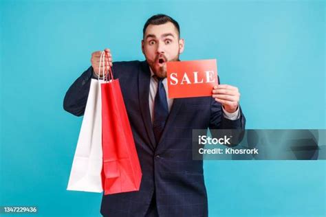 젊은 성인은 쇼핑백과 카드로 포즈를 취한 어두운 양복을 입고 충격을 받은 표정으로 카메라를 바라보며 깜짝 놀랐다 25 29세에