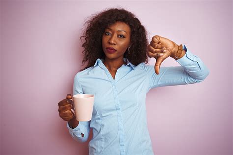 화가 얼굴 엄지 손가락으로 싫어하는 것을 보여주는 부정적인 기호 거부 개념으로 고립 된 분홍색 배경 위에 커피 컵을 마시는 젊은 아프리카 계 미국인 여성 개념에 대한 스톡