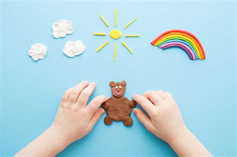 모델링 점토에서 갈색 곰을 만드는 아기 손 밝은 파란색 테이블 배경에 다채로운 무지개 태양과 흰색 구름 파스텔 색상 근접 촬영