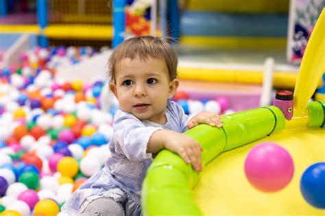 드레스를 입은 귀여운 유아 소녀가 무서운 표정으로 옆으로 올라갑니다 배경에 데이 어린이 케어 룸 놀이 센터의 거품 수영장입니다 고여 있는 물에 대한 스톡 사진 및 기타