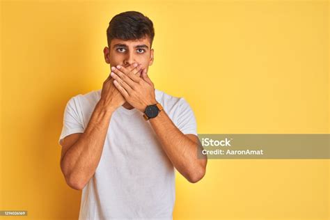 고립 된 노란색 배경 위에 서있는 흰색 티셔츠를 입은 젊은 인도 남자는 실수로 손으로 입을 덮는 충격을 받았습니다 비밀