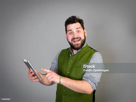 검은 머리와 전통적인 의상을 입은 수염을 가진 젊은 남자가 회색 배경 앞에 서서 태블릿에서 작동합니다 고급에 대한 스톡 사진 및