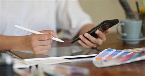 그래픽 디자이너가 스마트폰과 스타일러스 펜을 들고 나무 작업 책상 위에 놓고 커피 컵 컬러 가이드 연필꽂이로 둘러싸인 컴퓨터 태블릿에 그림을 그리는 동안 자른 이미지