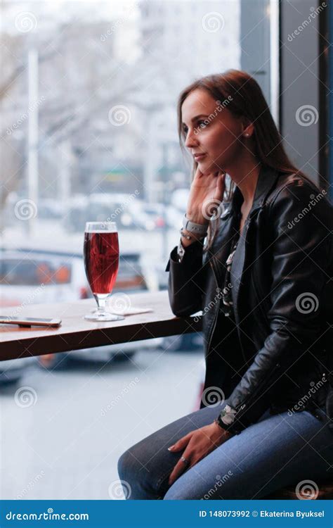 A Young Beautiful Woman Is Sitting On A Bar Stool By A Large Window