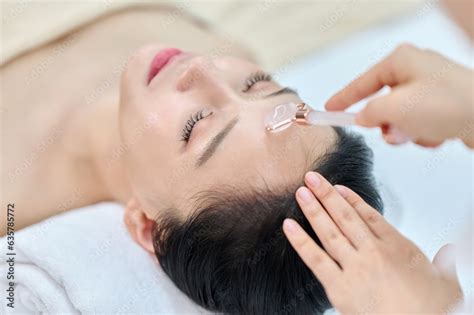 아시아 한국의 젊은 여성이 스파 또는 피부관리전문점 또는 피부과 병원에서 침대에 누워 베이지색 대형 타올을 덮고 눈을 감고 있고 전문 피부관리사가 마사지롤러를 들고 이마를
