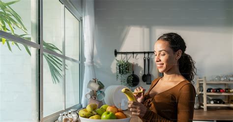 젊은 매력적인 라틴계 여성은 집에서 부엌에서 과일을 먹습니다 행복한 얼굴을 가진 아름다운 활동적인 여성 스탠드 바나나 먹기 건강 관리 웰빙을위한 건강 식품을 집에서